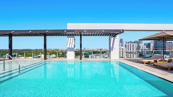 Uma piscina no terraço com água azul cristalina, uma pérgula, cadeiras de descanso e vista para o horizonte da cidade.
