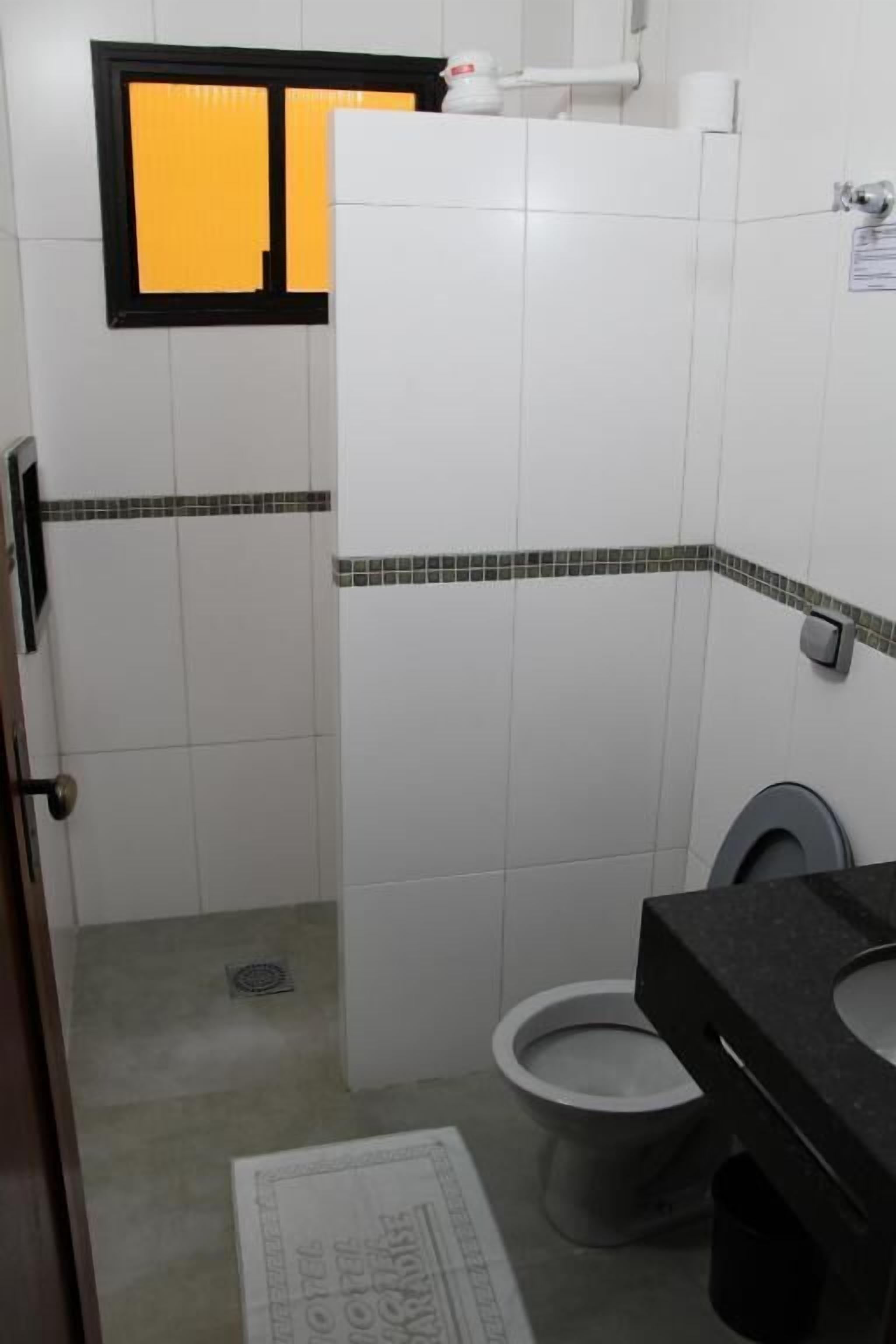 Casa de banho com sanita, bidé e lavatório. Tem uma janela com estrutura preta e uma persiana amarela. As paredes estão revestidas com azulejos brancos e verdes.