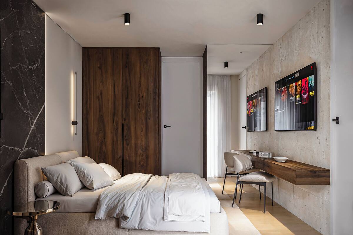 Quarto moderno com cama grande, guarda-roupa de madeira, escrivaninha com cadeira e televisão na parede.
