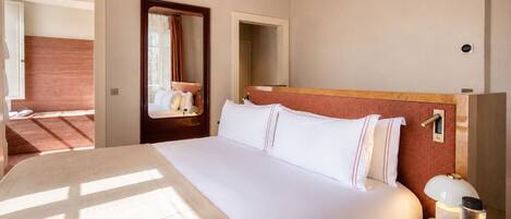 Quarto de hotel com uma cama grande, uma mesa de cabeceira com abajur, um espelho e uma porta que dá para outro cômodo.