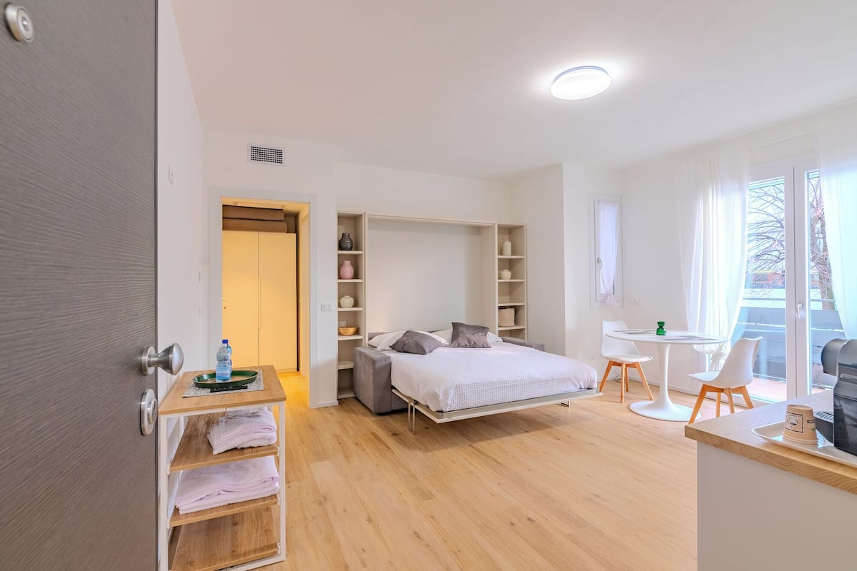 Quarto de hotel moderno com cama, pequena mesa de trabalho, cadeira, guarda-roupa e banheiro.