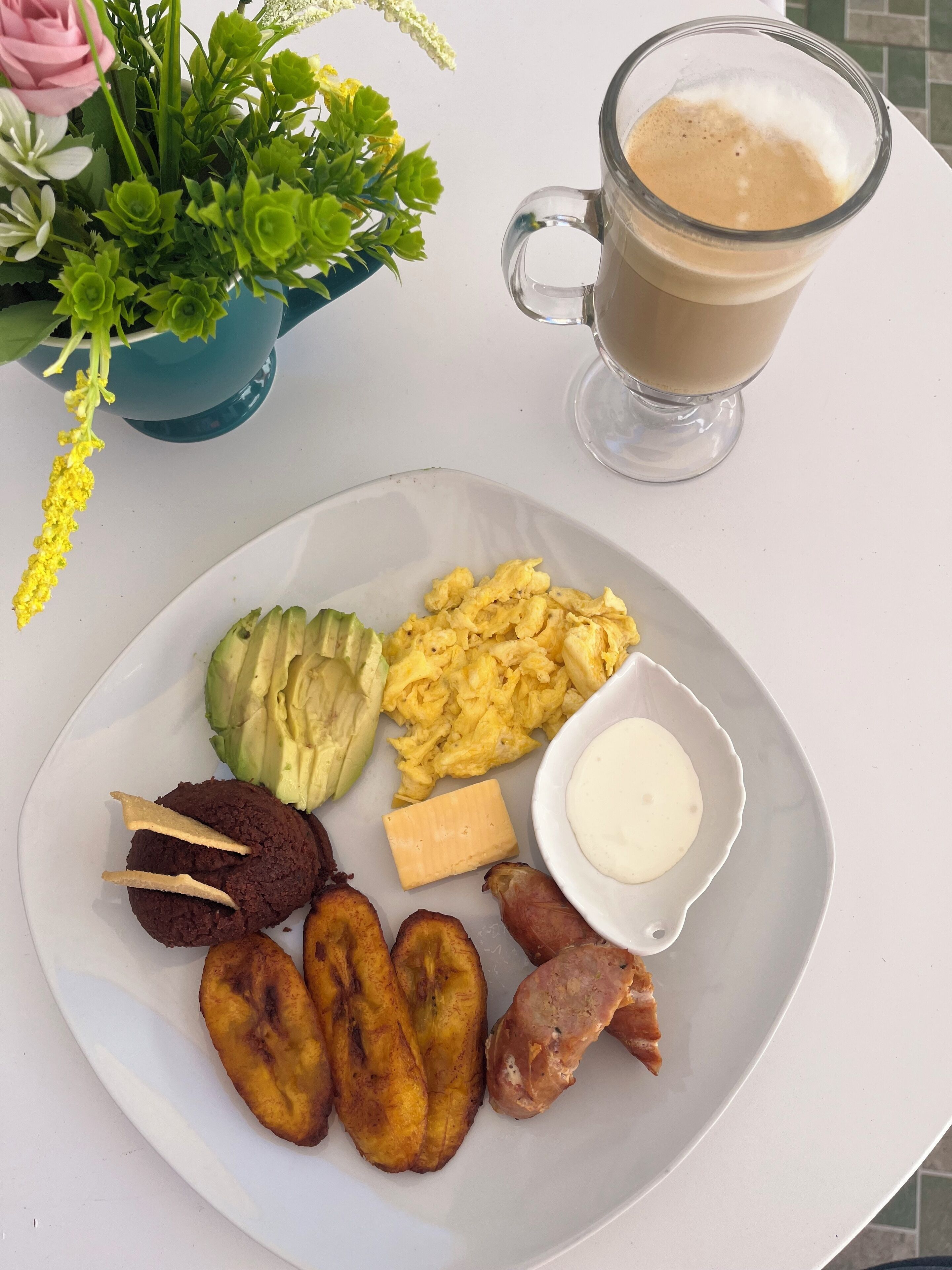 Prato de café da manhã com ovos mexidos, abacate, queijo, uma pequena tigela de manteiga, plantains fritos e uma xícara de café.