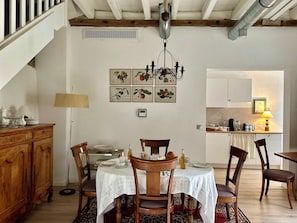 Uma sala de jantar com uma mesa de madeira posta para quatro pessoas, rodeada por cadeiras. Há um lustre sobre a mesa, e uma cozinha é visível ao fundo.