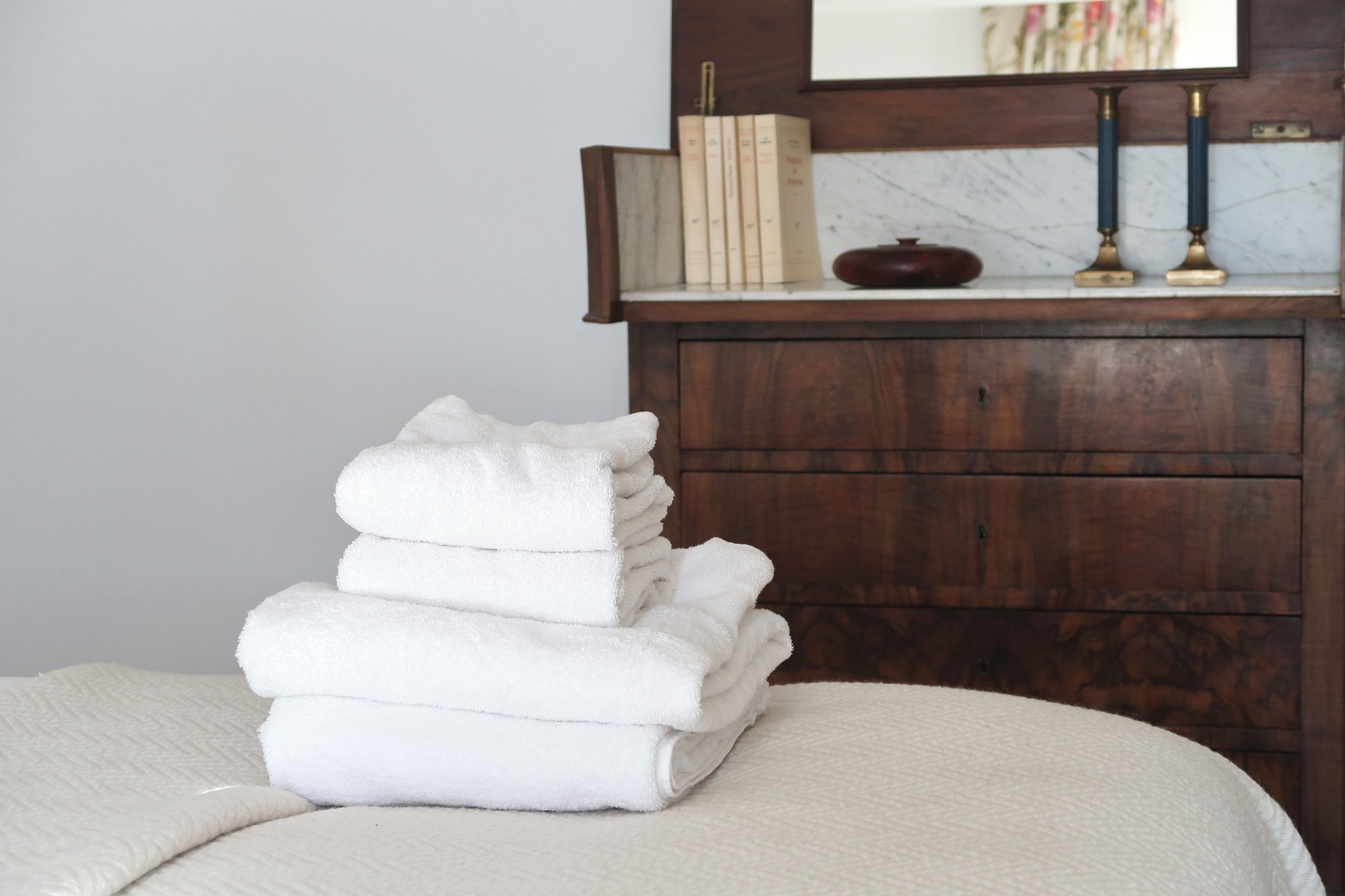 Uma pilha de toalhas brancas em uma cama, com uma cômoda de tampo de mármore ao fundo.