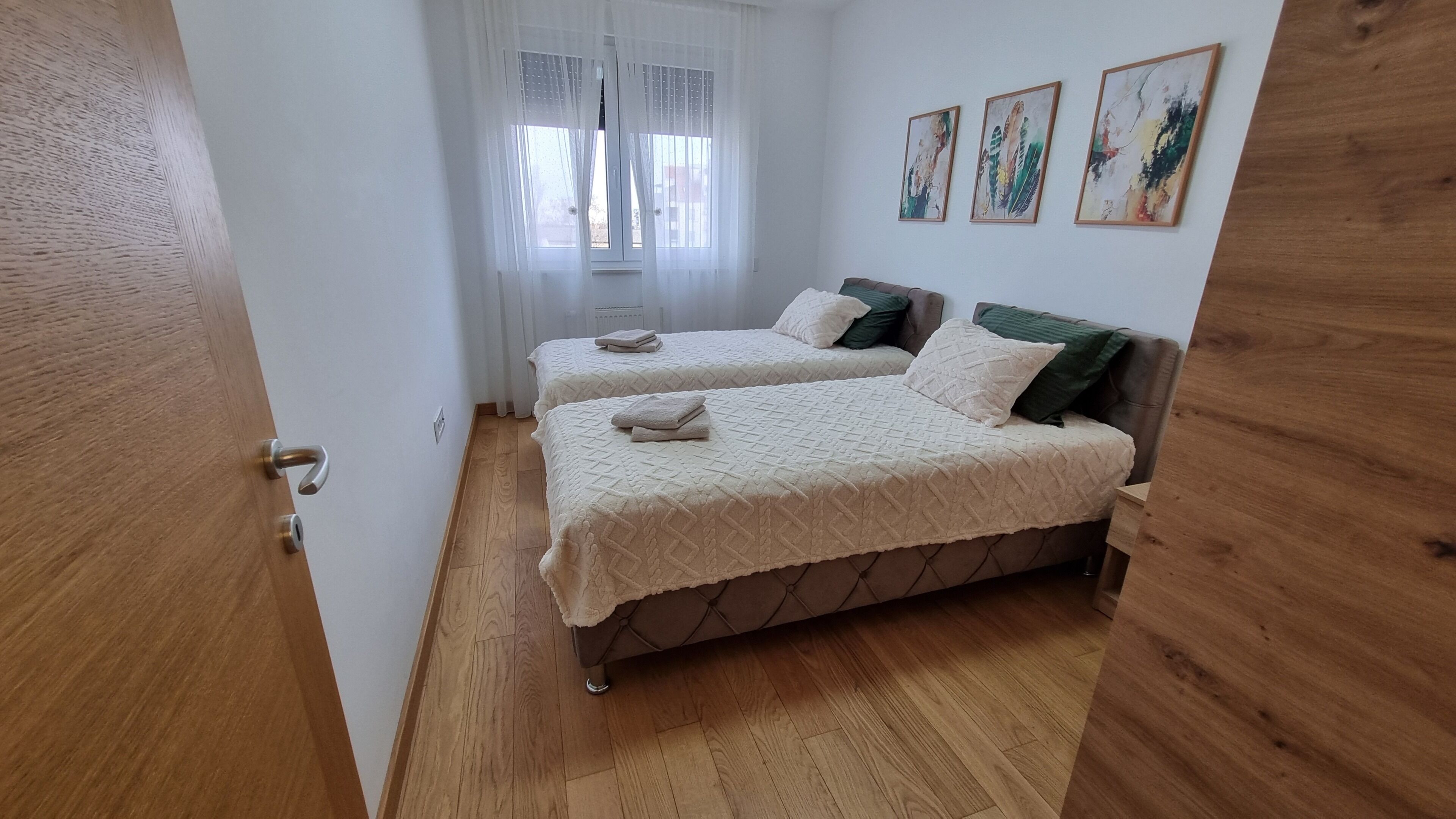 Quarto com duas camas, soalho de madeira, janela com cortinas leves e três obras de arte fixadas na parede.
