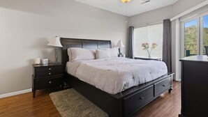 Quarto com cama, mesas de cabeceira, cômoda, ventoinha no teto e janela com cortinas.