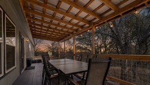 Um deck externo com mesa e cadeiras de madeira, cercado por um corrimão de madeira e vista para árvores.