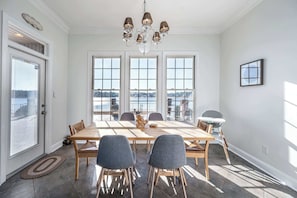 Uma sala de jantar com uma mesa grande, cadeiras, um candelabro e janelas amplas com vista.