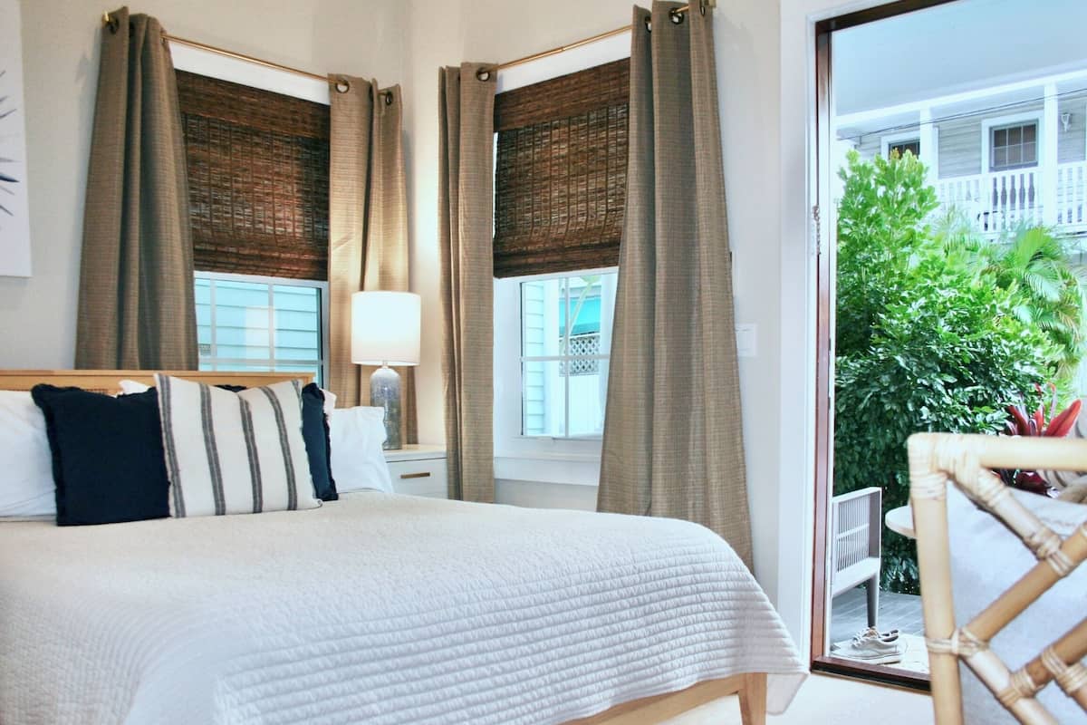 Quarto com cama, cortinas, mesa de cabeceira, abajur e vista para uma varanda com plantas.