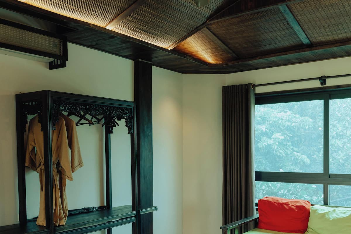 Quarto com cama, ventilador de teto, janela com cortinas e prateleira de madeira com roupas.