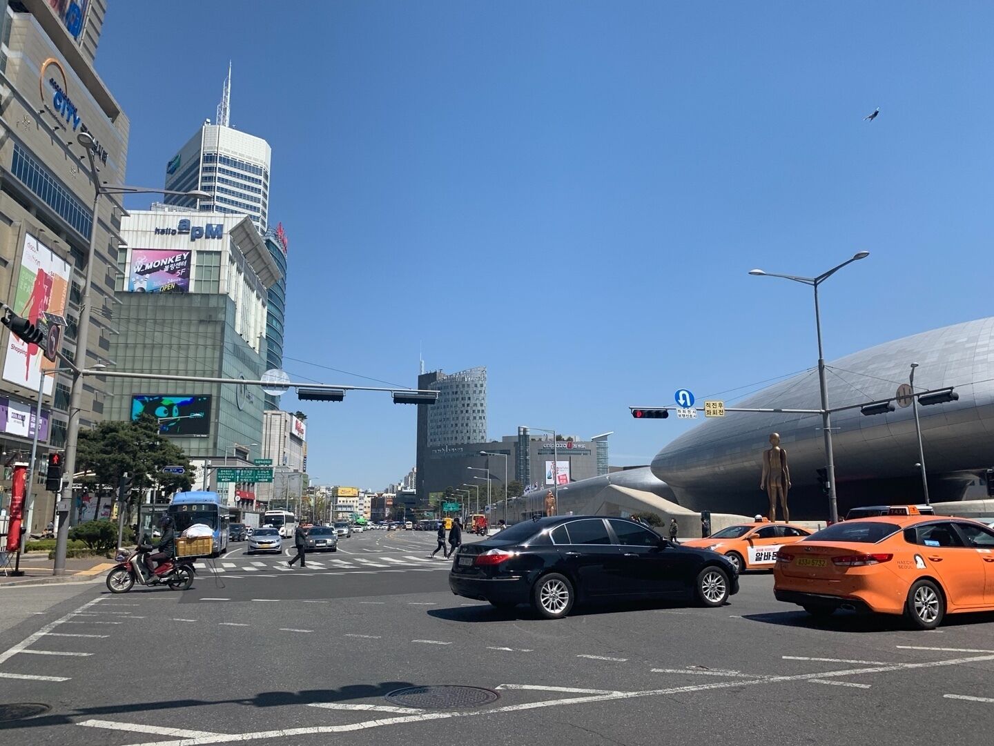 Uma rua urbana movimentada com edifícios modernos, uma grande estrutura esférica e diversos veículos, incluindo carros e um ônibus.