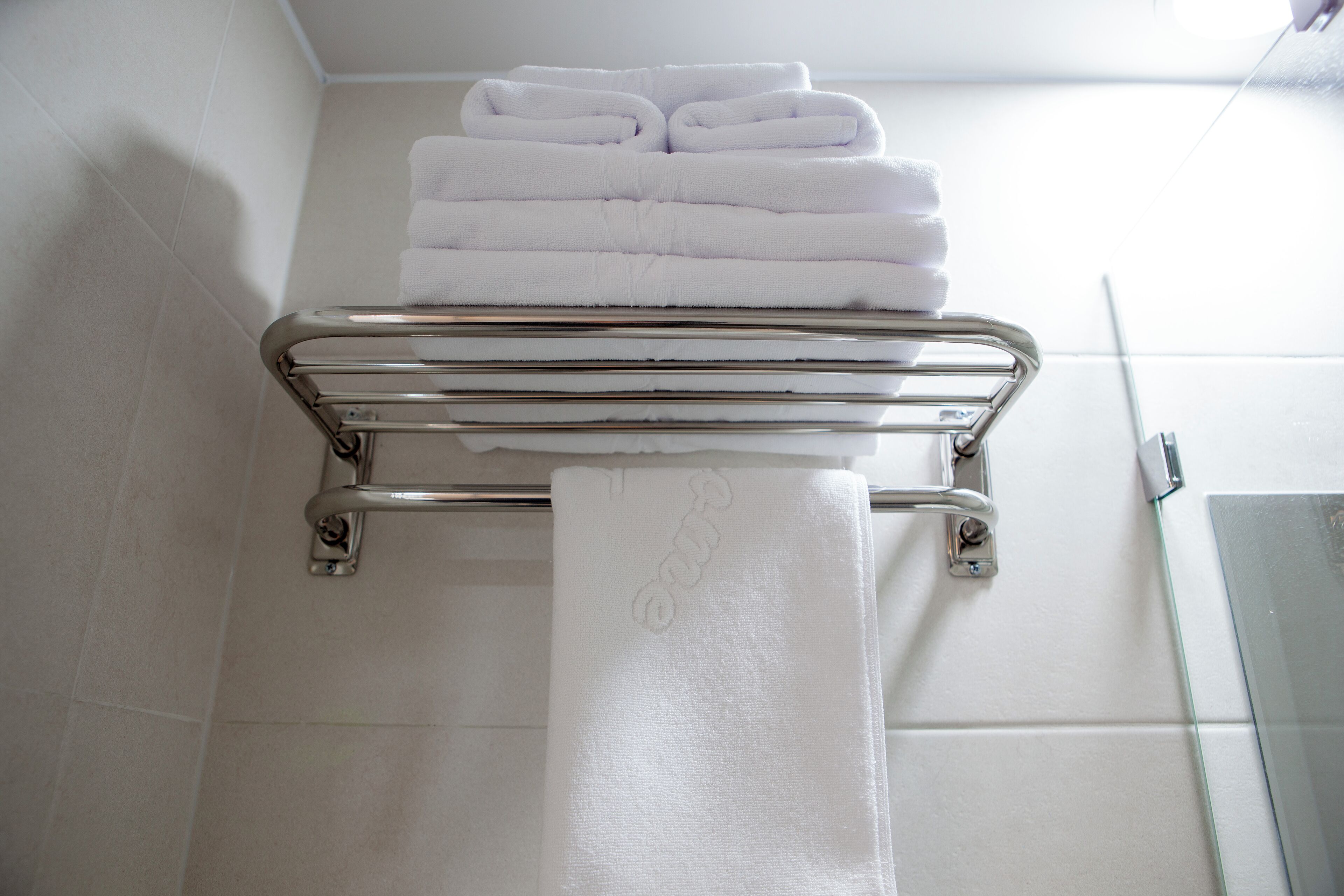 Uma pilha de toalhas brancas dobradas em um porta-toalhas de metal cromado em um banheiro com azulejos brancos.