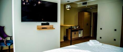 Quarto de hotel com cama, televisão na parede, escrivaninha com candeeiro e guarda-roupa.