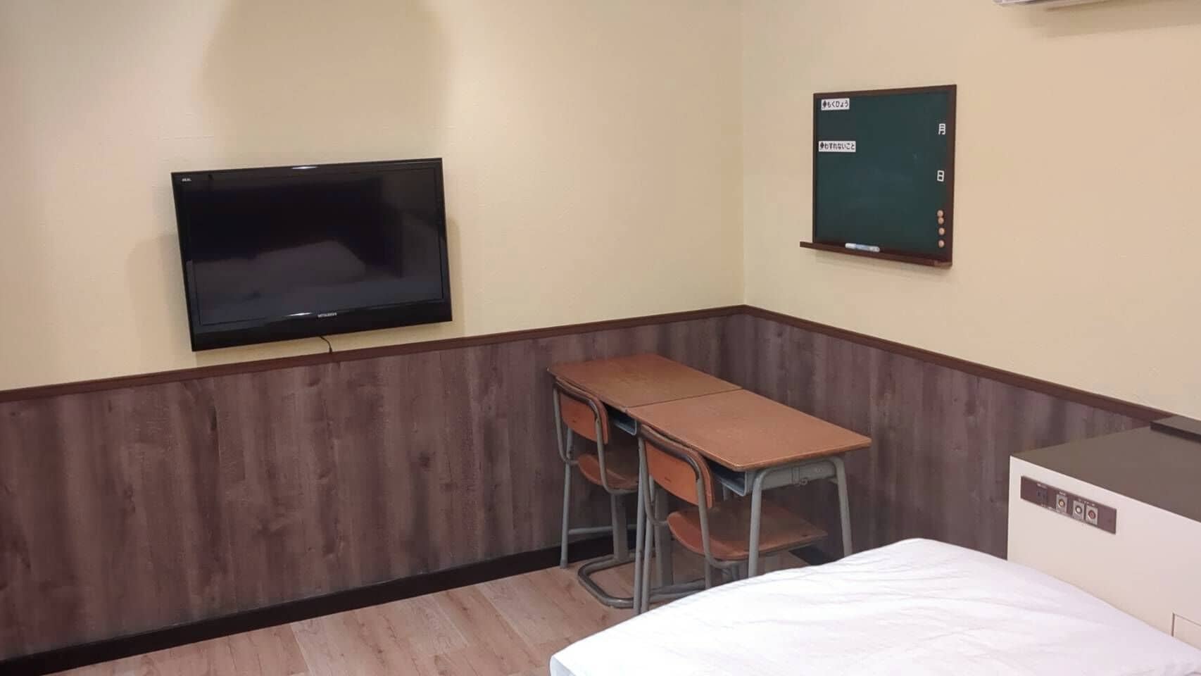 Um quarto com uma cama, uma escrivaninha com cadeiras, uma televisão e um quadro negro.