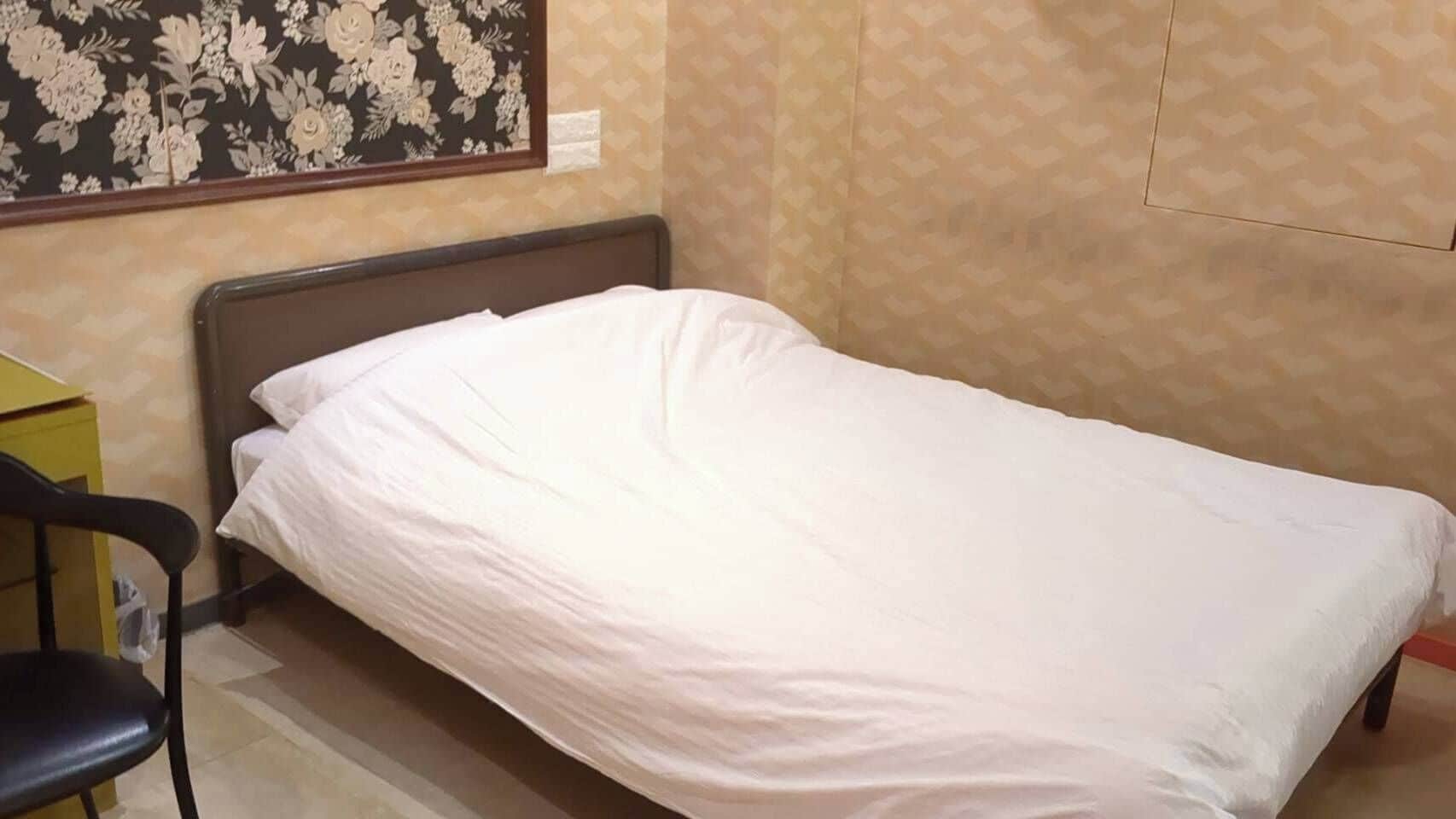 Uma cama de solteiro com roupa de cama branca em um quarto com papel de parede estampado e uma pequena escrivaninha.