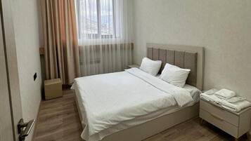 Quarto de hotel bem arrumado, com uma cama, uma mesa de cabeceira com abajur e uma janela com cortinas.