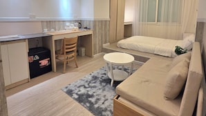Quarto de hotel compacto com cama, escrivaninha, cadeira, mesa redonda pequena e ventilador de teto.