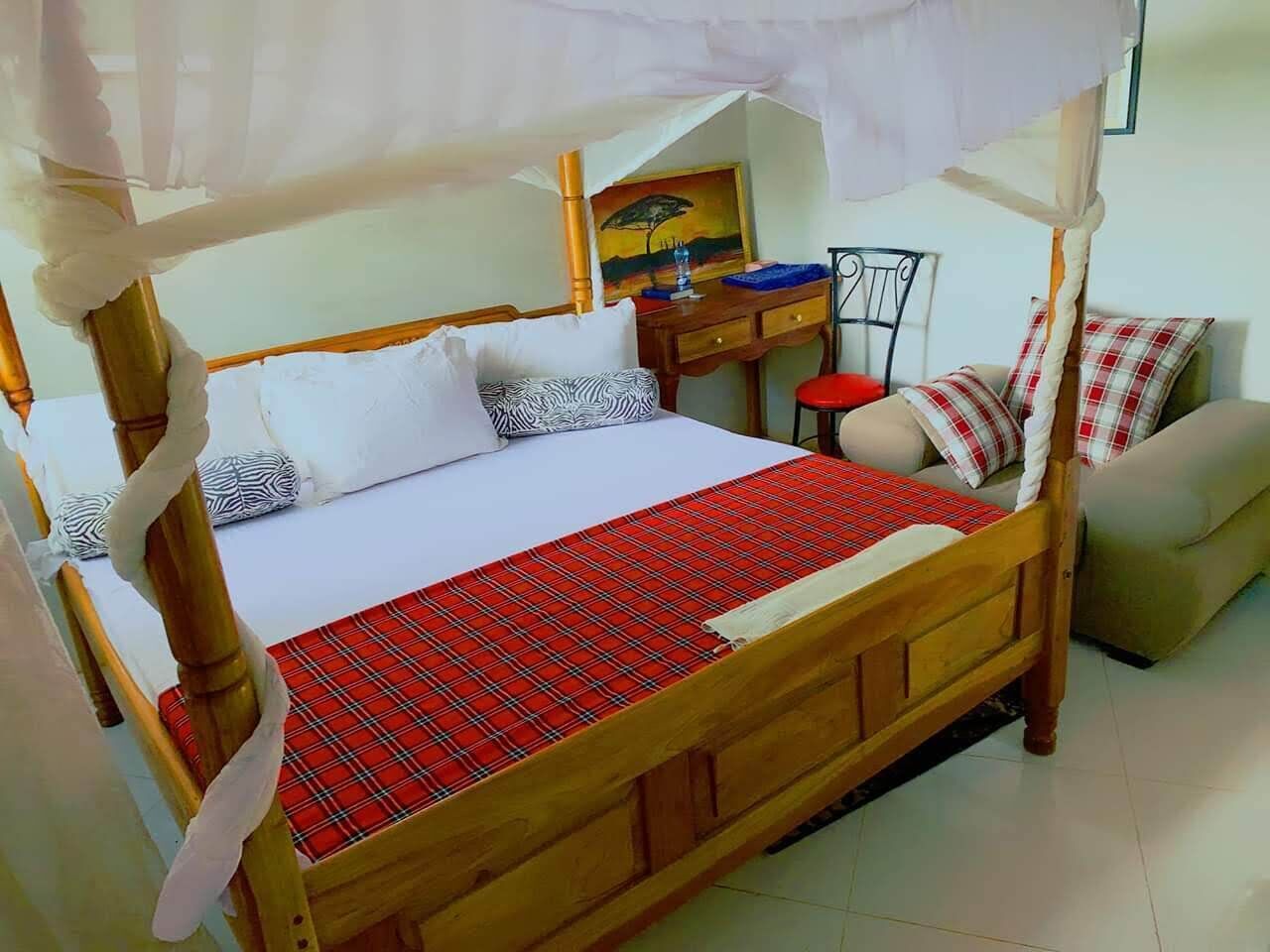 Uma cama de madeira com colcha xadrez vermelha e branca, uma mesa de cabeceira de madeira, uma cadeira e um sofá com almofadas estampadas.