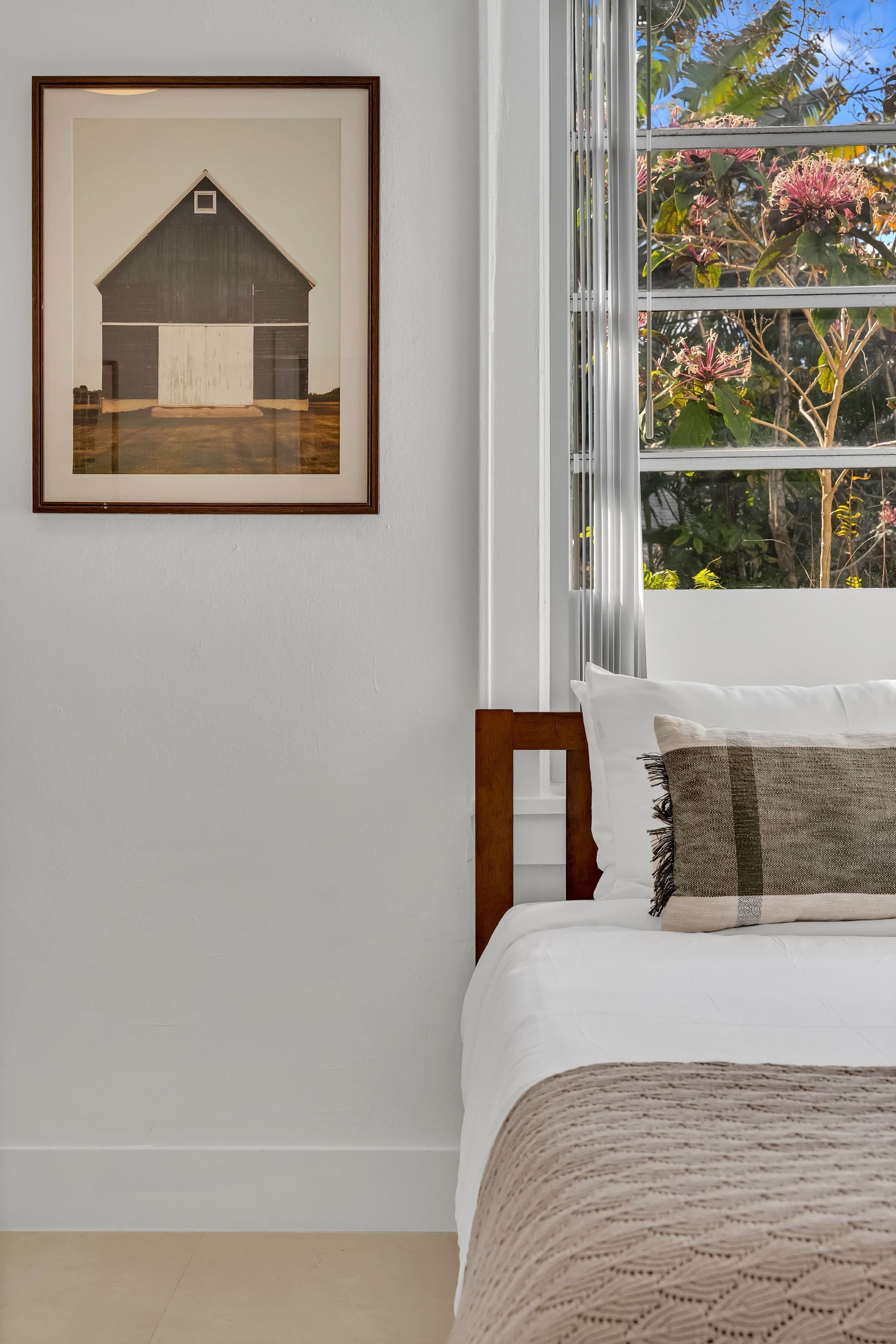 Quarto com cama, almofadas, quadro na parede e janela com vista para árvores e flores.