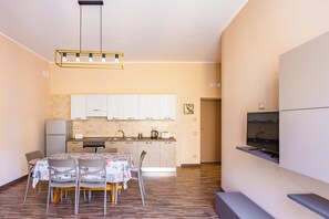 Uma cozinha moderna com armários brancos, forno embutido e micro-ondas. Há uma área de jantar com mesa e cadeiras, e uma televisão fixada na parede.