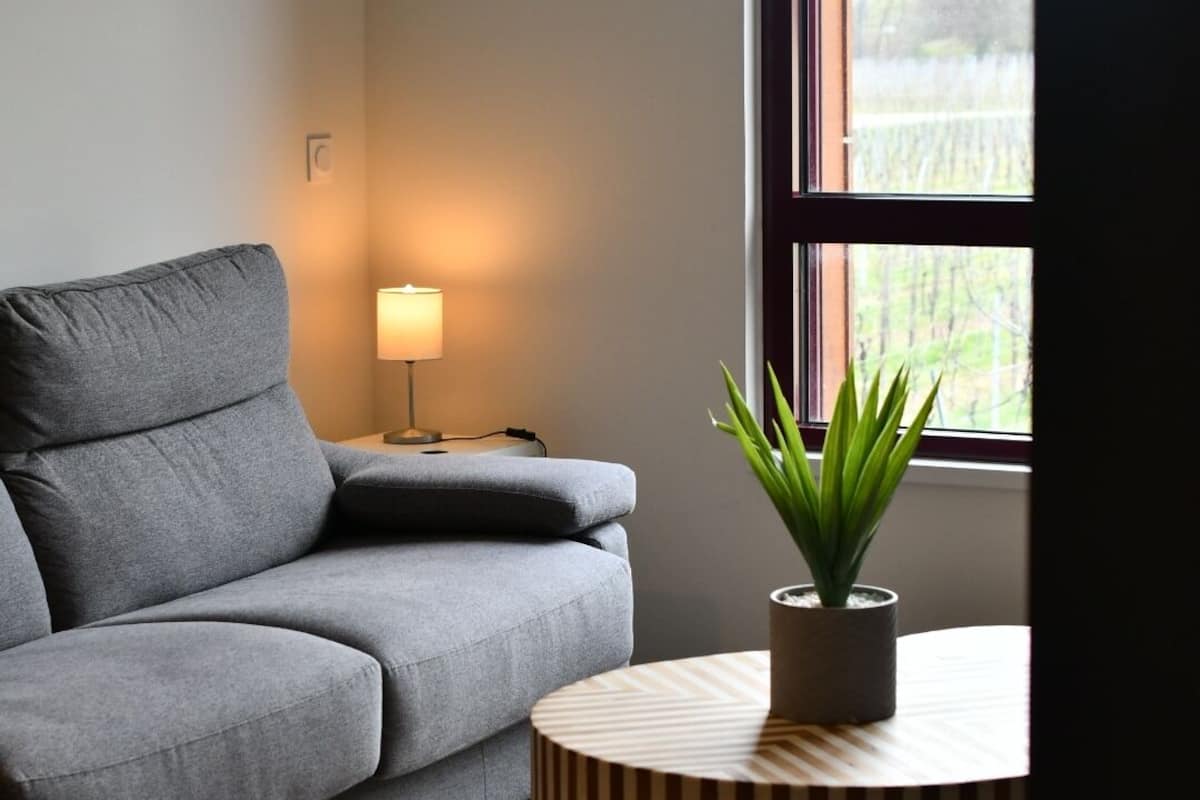Uma sala de estar moderna com um sofá cinza, uma mesa de apoio redonda de madeira, um abajur e um vaso de planta perto de uma janela.
