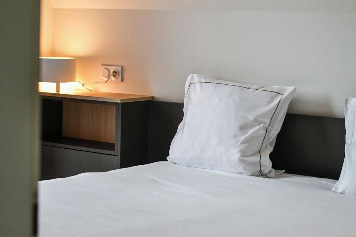 Uma cama bem arrumada com lençóis brancos, uma mesa de cabeceira com abajur e uma cabeceira escura.
