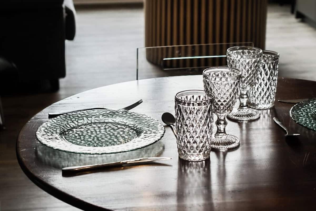 Uma mesa de jantar com copos, um prato estampado, uma planta em vaso e uma janela com vista para edifícios.