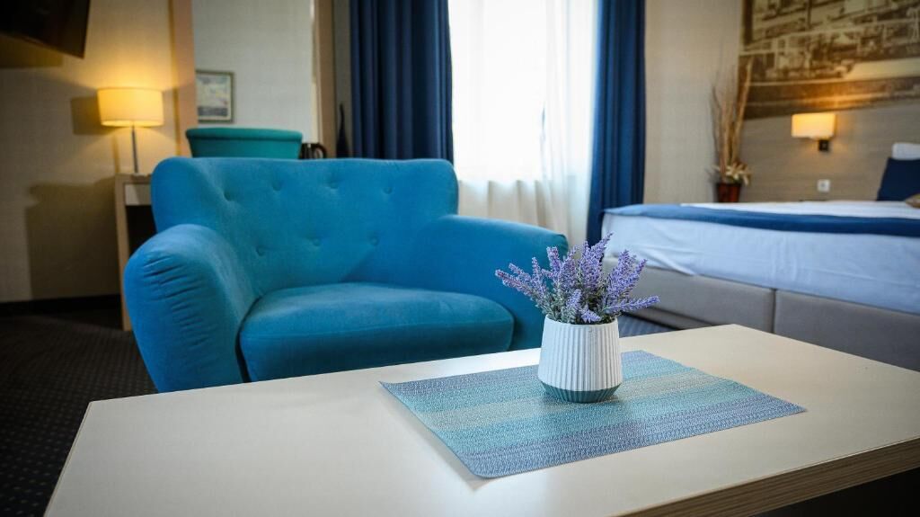 Quarto de hotel com um sofá azul, uma mesa branca com um vaso de flores, uma cama com cobre azul e uma luminária na parede.