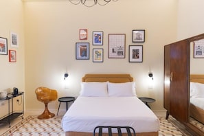 Quarto de hotel com cama, guarda-roupa de madeira, secretária com cadeira, mesa redonda pequena e uma cadeira. Quadros emoldurados na parede.