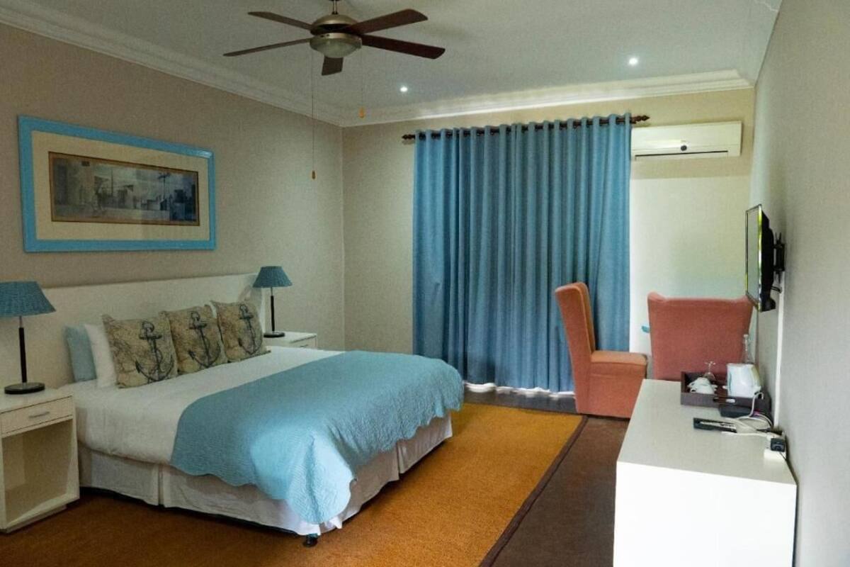 Quarto de hotel com cama, escrivaninha, cadeira, ventoinha no teto e janela com cortinas.