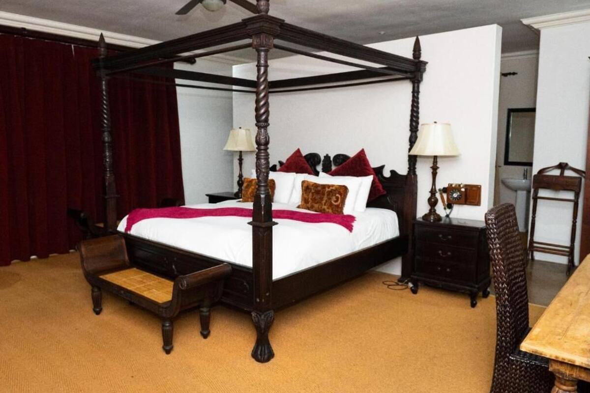 Uma cama com dossel e cortina, um criado-mudo de madeira, uma cadeira e uma mesa em um quarto de hotel.