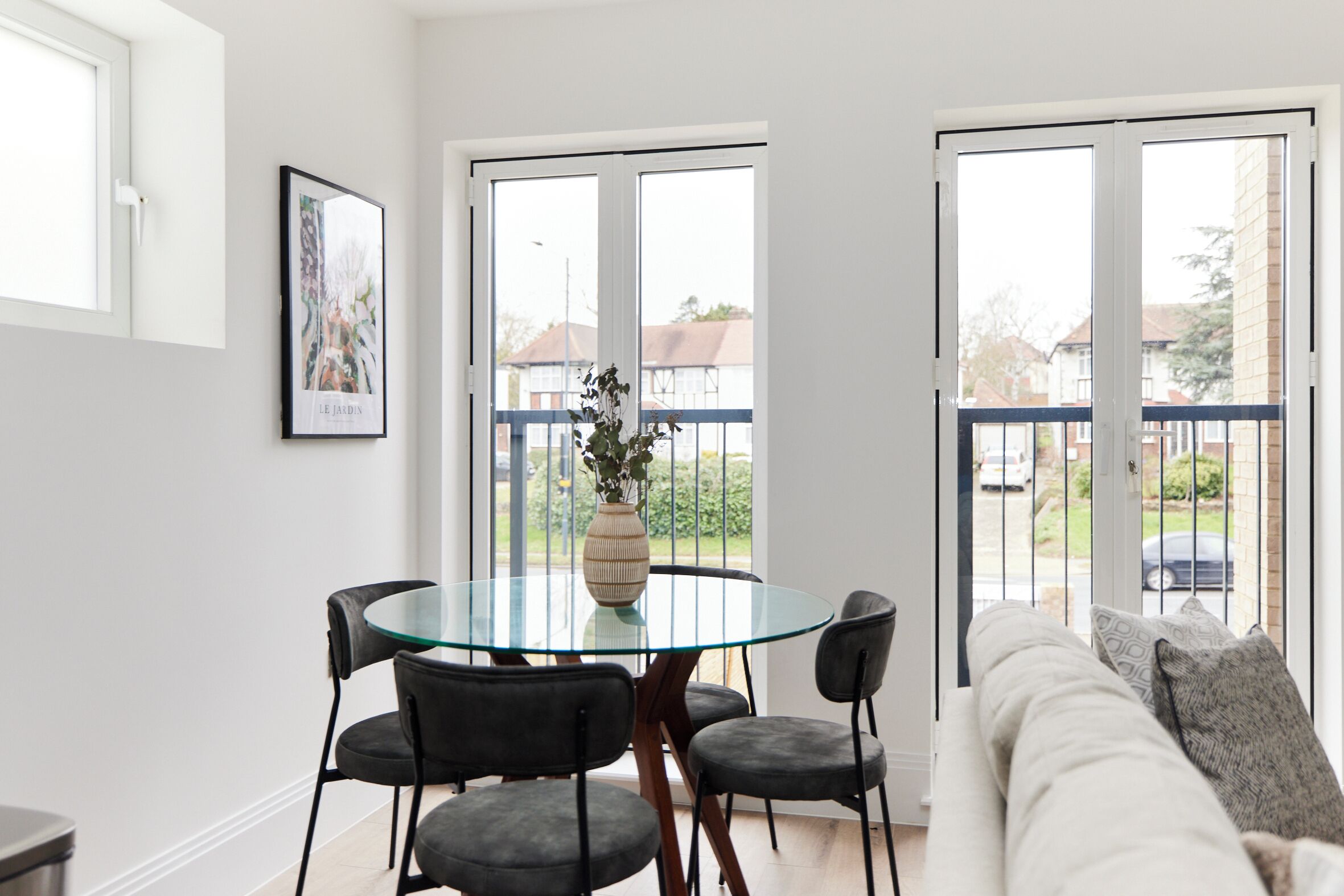 Uma área de refeições moderna com mesa de vidro e quatro cadeiras, um sofá branco com almofadas e uma janela grande com vista para casas e um carro.