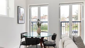 Uma área de refeições moderna com mesa de vidro e quatro cadeiras, um sofá branco com almofadas e uma janela grande com vista para casas e um carro.