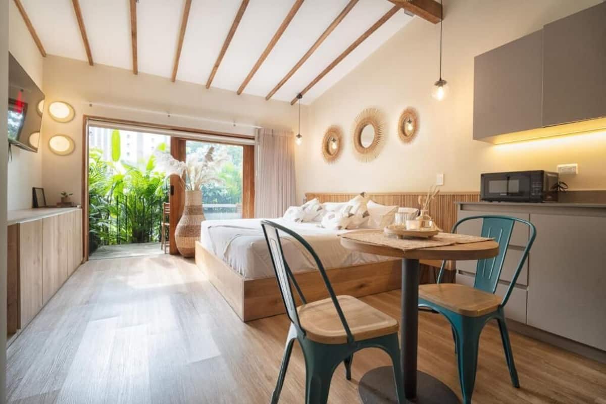 Quarto moderno com cama, mesa de jantar e cadeiras. Há uma área de cozinha com micro-ondas e armários. O quarto tem piso de madeira e vista para o verde lá fora.