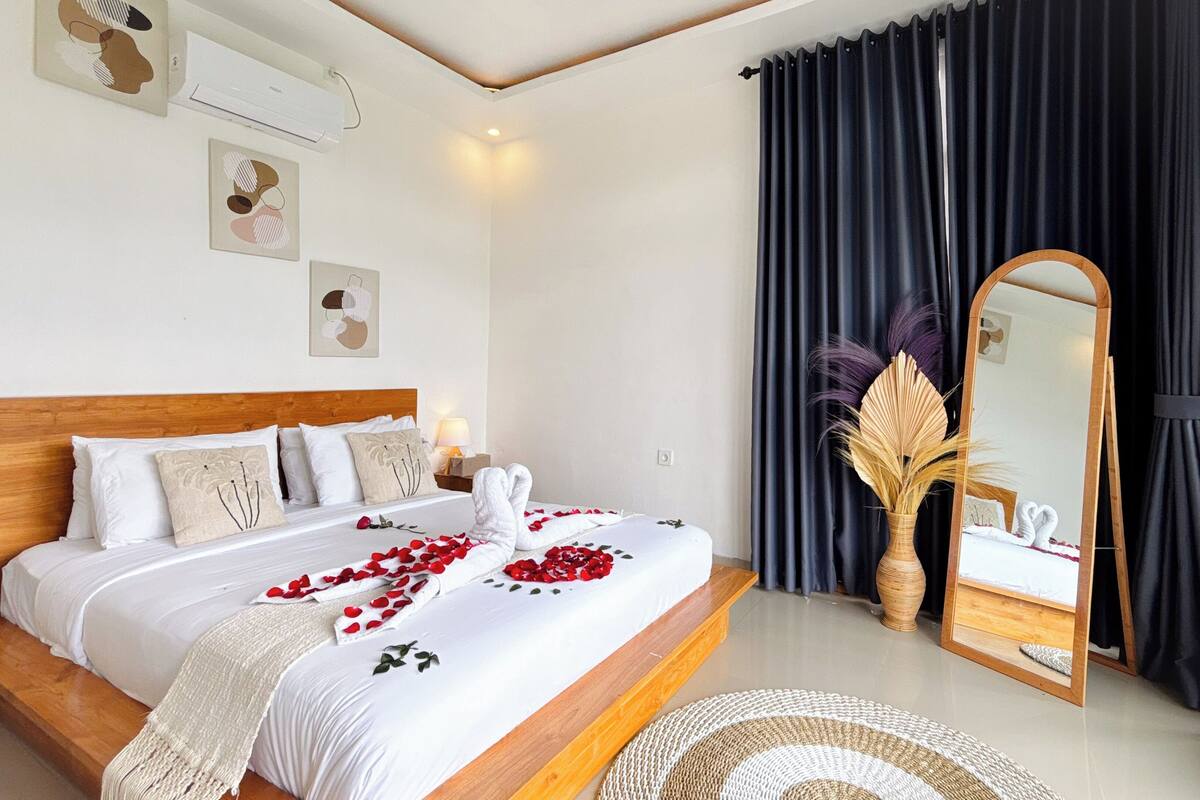 Quarto de hotel moderno com cabeceira de madeira, cama grande com roupa de cama branca, espelho e vista para o exterior.