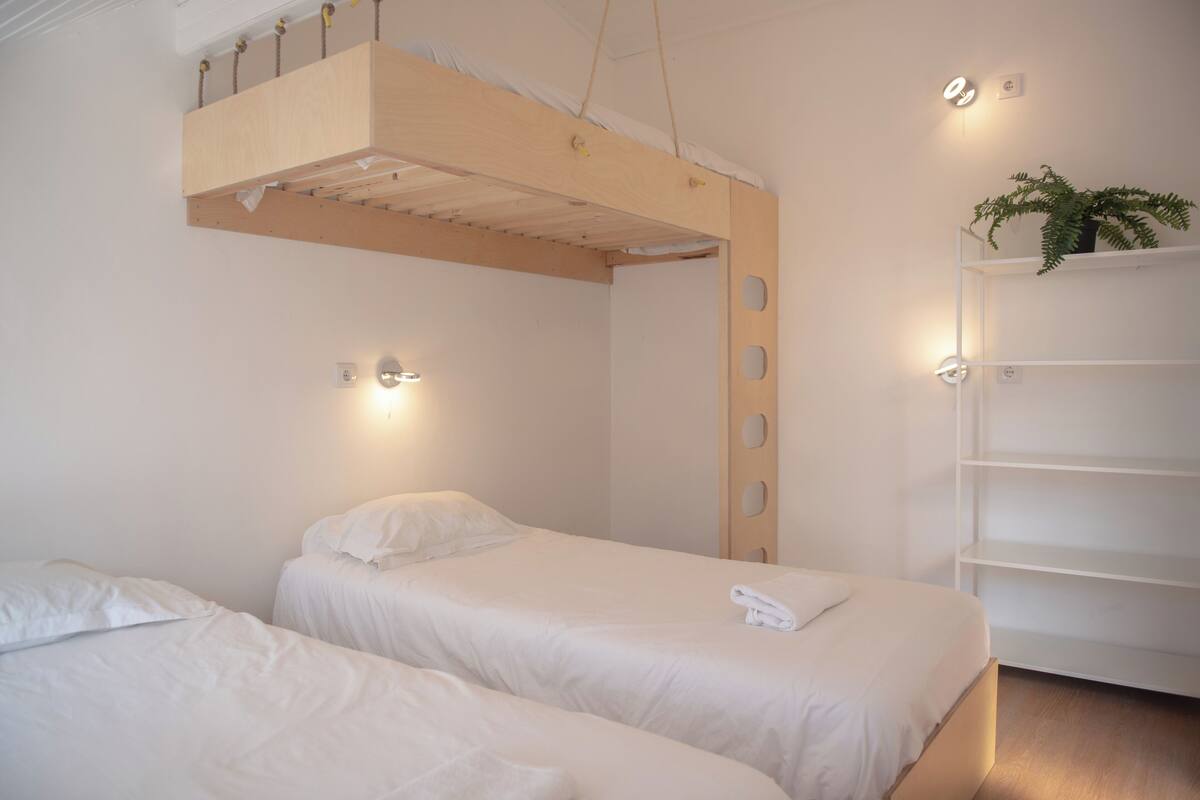 Quarto com duas camas de solteiro, uma cama beliche, um guarda-roupa branco e um vaso de plantas.