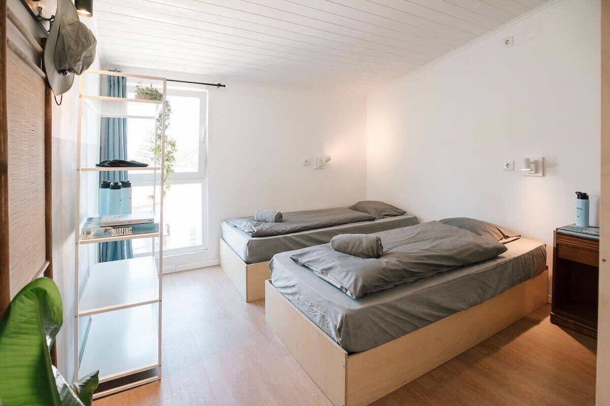Quarto minimalista com duas camas de solteiro, piso de madeira, janela com cortinas e uma estante com livros e uma planta.
