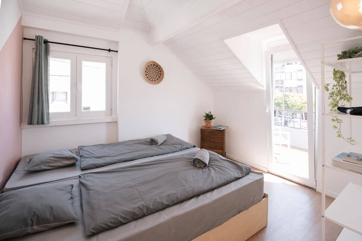 Quarto com duas camas, janela com cortinas, uma mesinha com uma planta e vista para o exterior.
