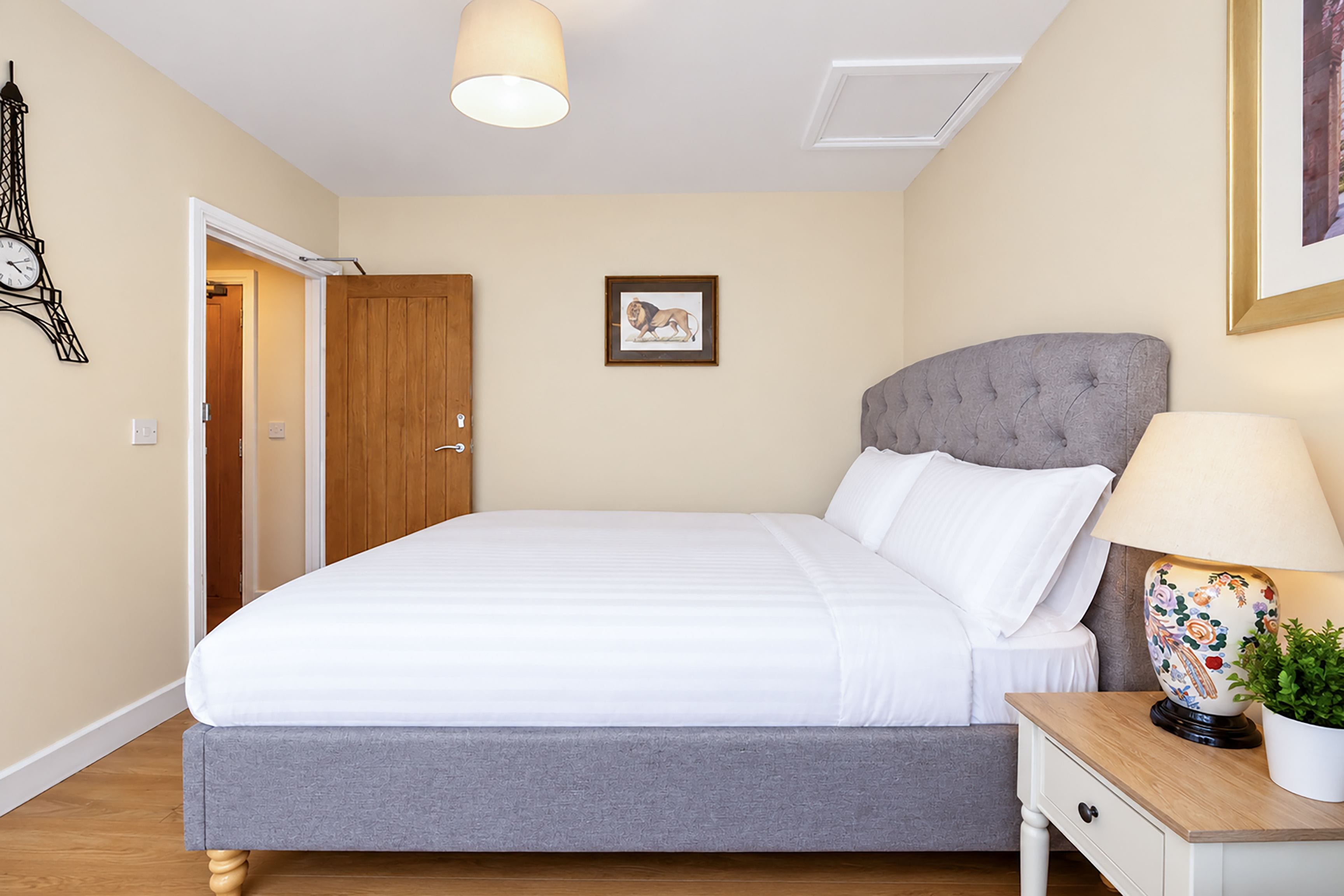 Quarto com cama, mesa de cabeceira, abajur e porta para outro cômodo.