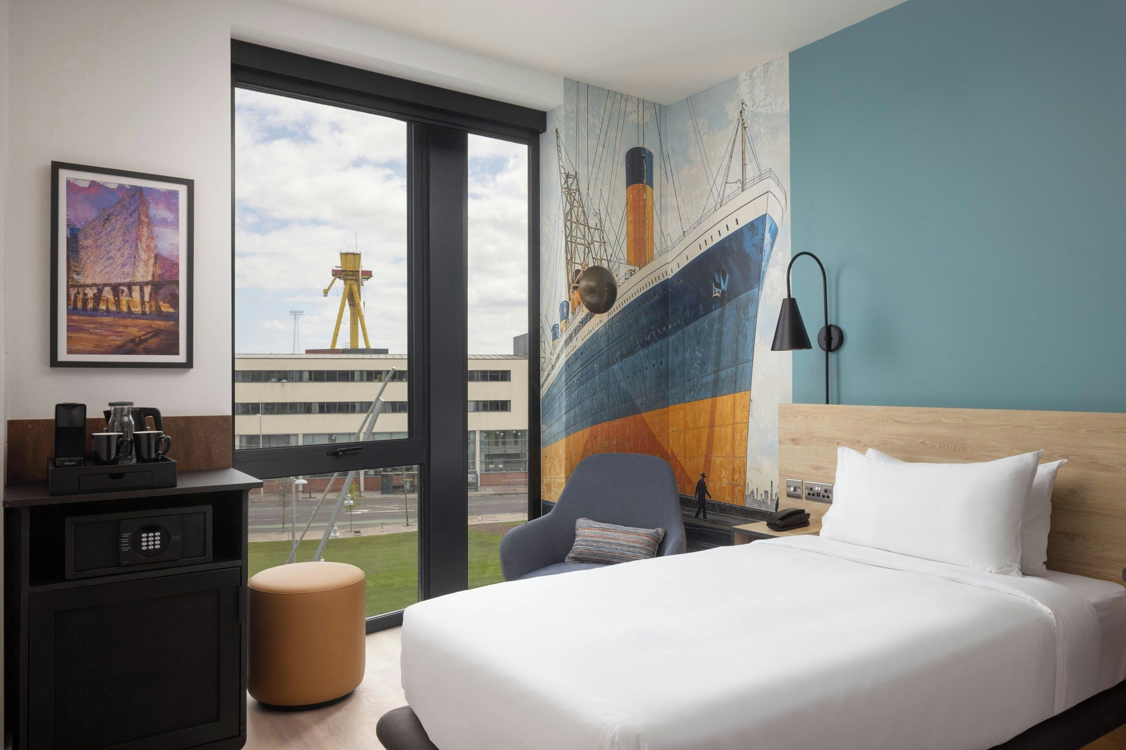 Quarto de hotel com uma cama grande, uma escrivaninha, uma cadeira, uma cafeteira, um abajur e um mural de um navio.