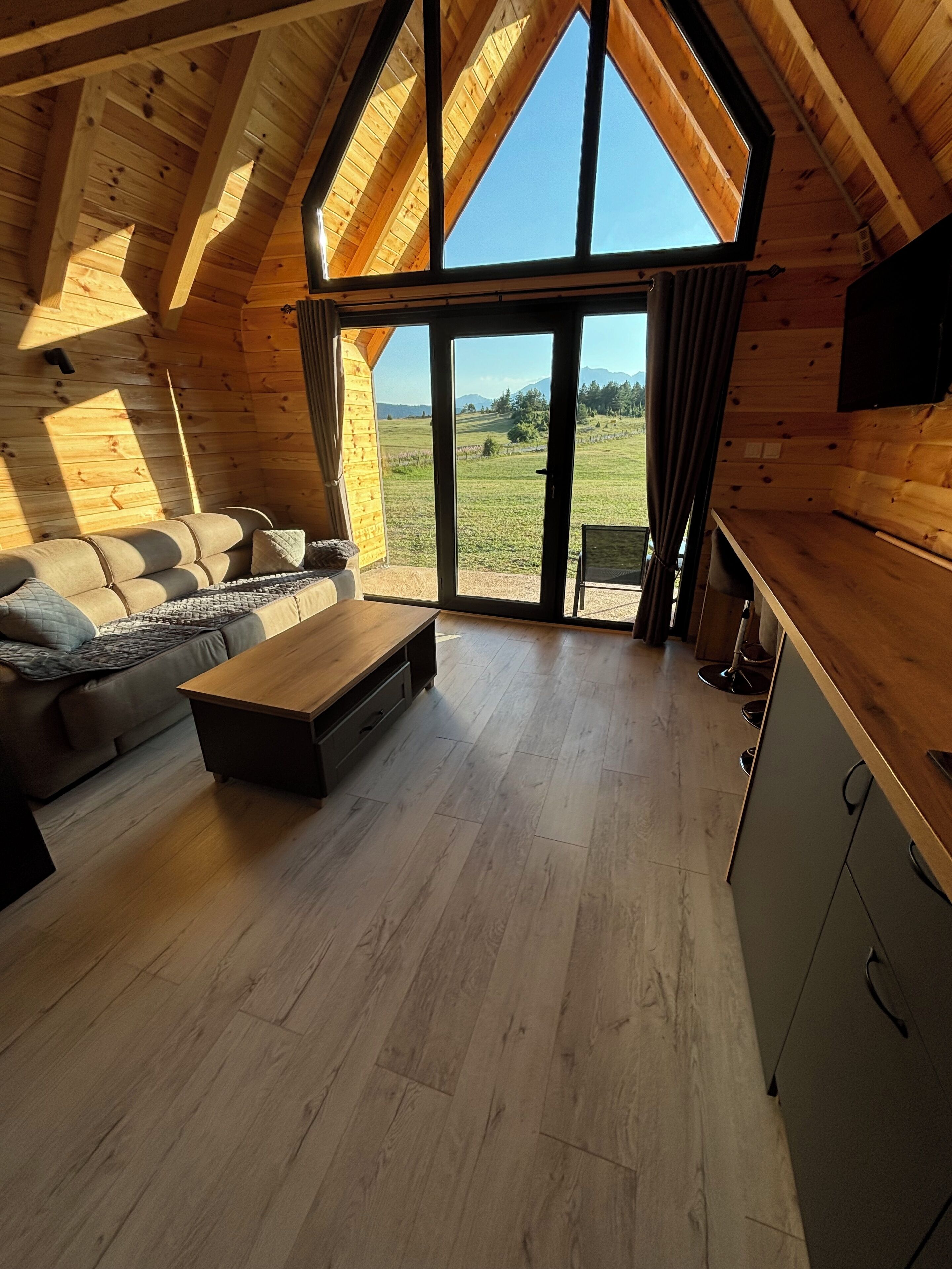 Interior acolhedor de uma cabana, com teto de madeira, um sofá, uma mesa de centro e vista para uma paisagem verde através de uma grande janela.