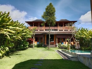 Um edifício de dois andares com varanda, rodeado por vegetação exuberante e uma piscina.