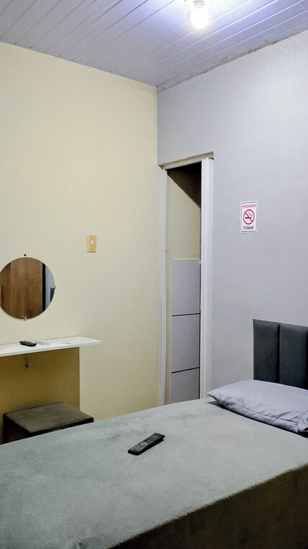 Quarto com cama, mesa de cabeceira, espelho, guarda-roupa e placa de 'Proibido Fumar'.