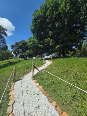 Uma trilha de cascalho ladeada por pedras, conduzindo a uma estrutura de madeira em meio a vegetação exuberante e árvores.