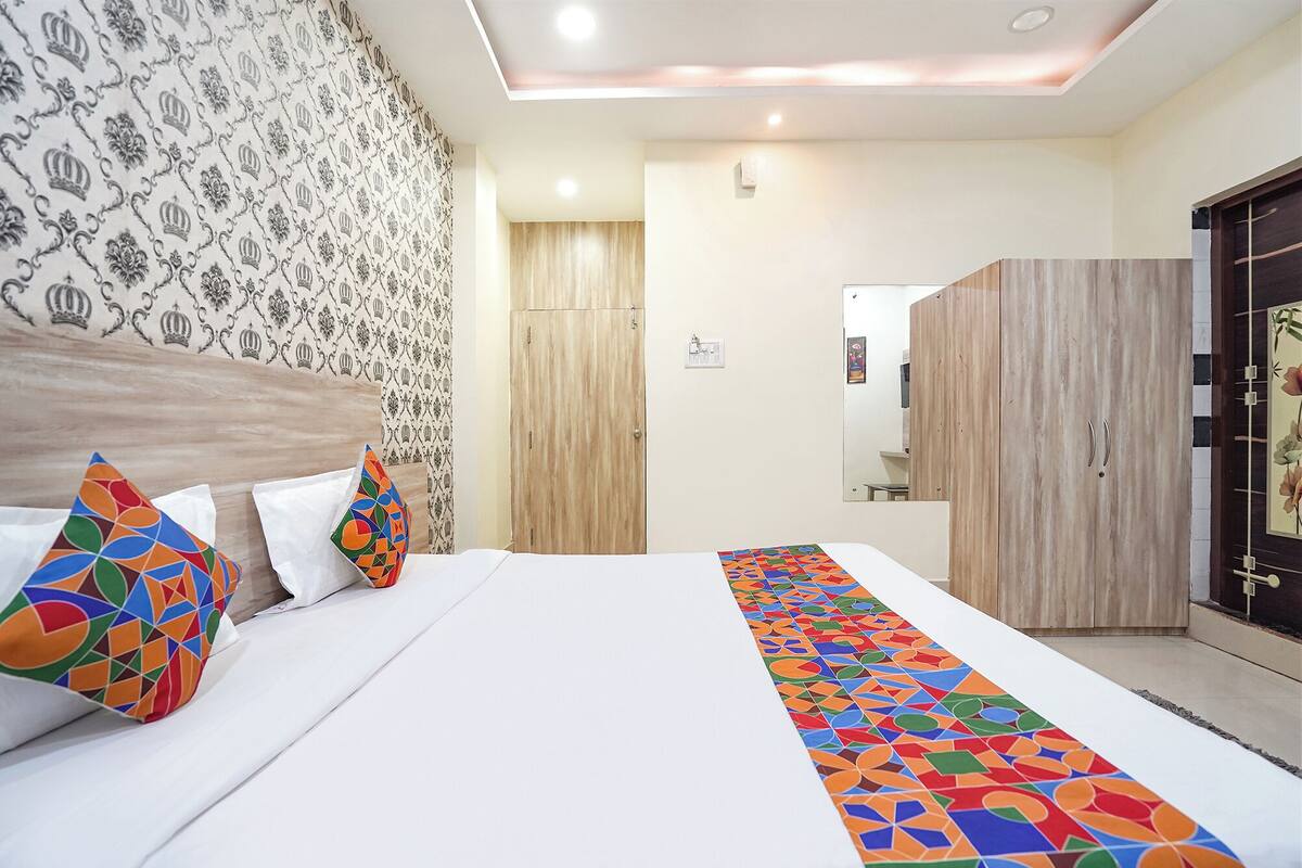 Quarto de hotel com uma cama grande, cabeceira de madeira, papel de parede estampado, cobre-leito colorido, guarda-roupa e um espelho.