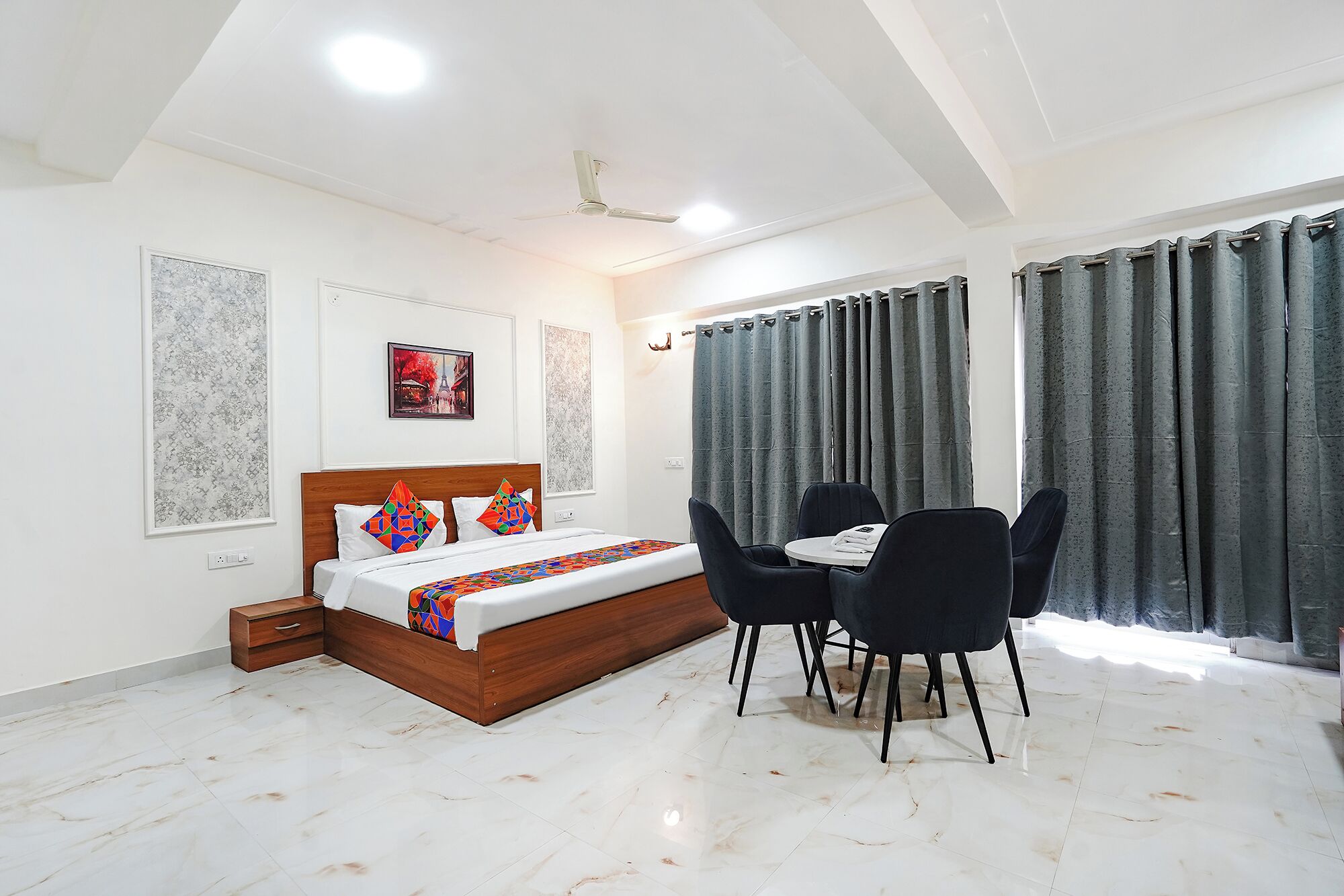 Quarto com cama, mesa de cabeceira, cadeira, mesinha e janela com cortinas.
