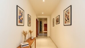 Um corredor com quadros de arte abstrata nas paredes, um banco de madeira com livros e objetos decorativos, e uma porta no final.