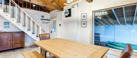 Uma mesa de jantar de madeira com bancos, uma escada com grades brancas e vista para um barco verde através de uma grande janela.