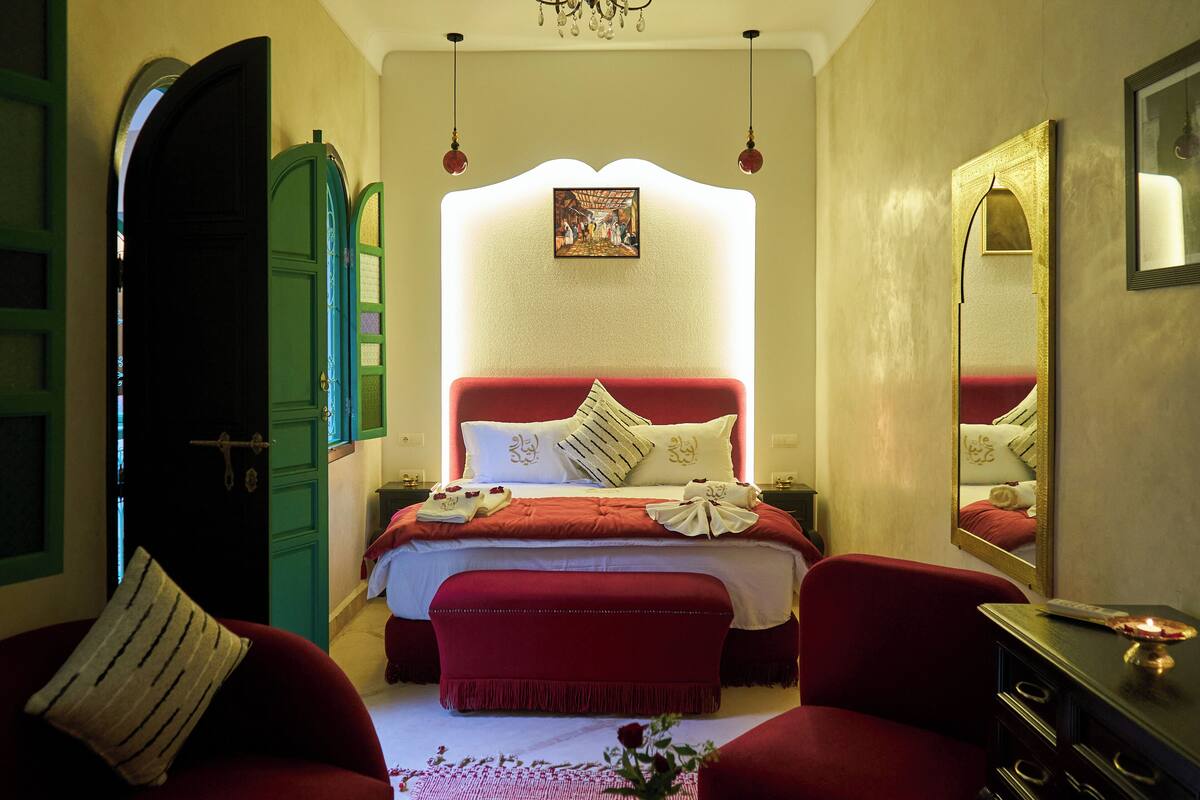 Quarto com cama, uma cadeira vermelha, um banco, um espelho e uma porta.