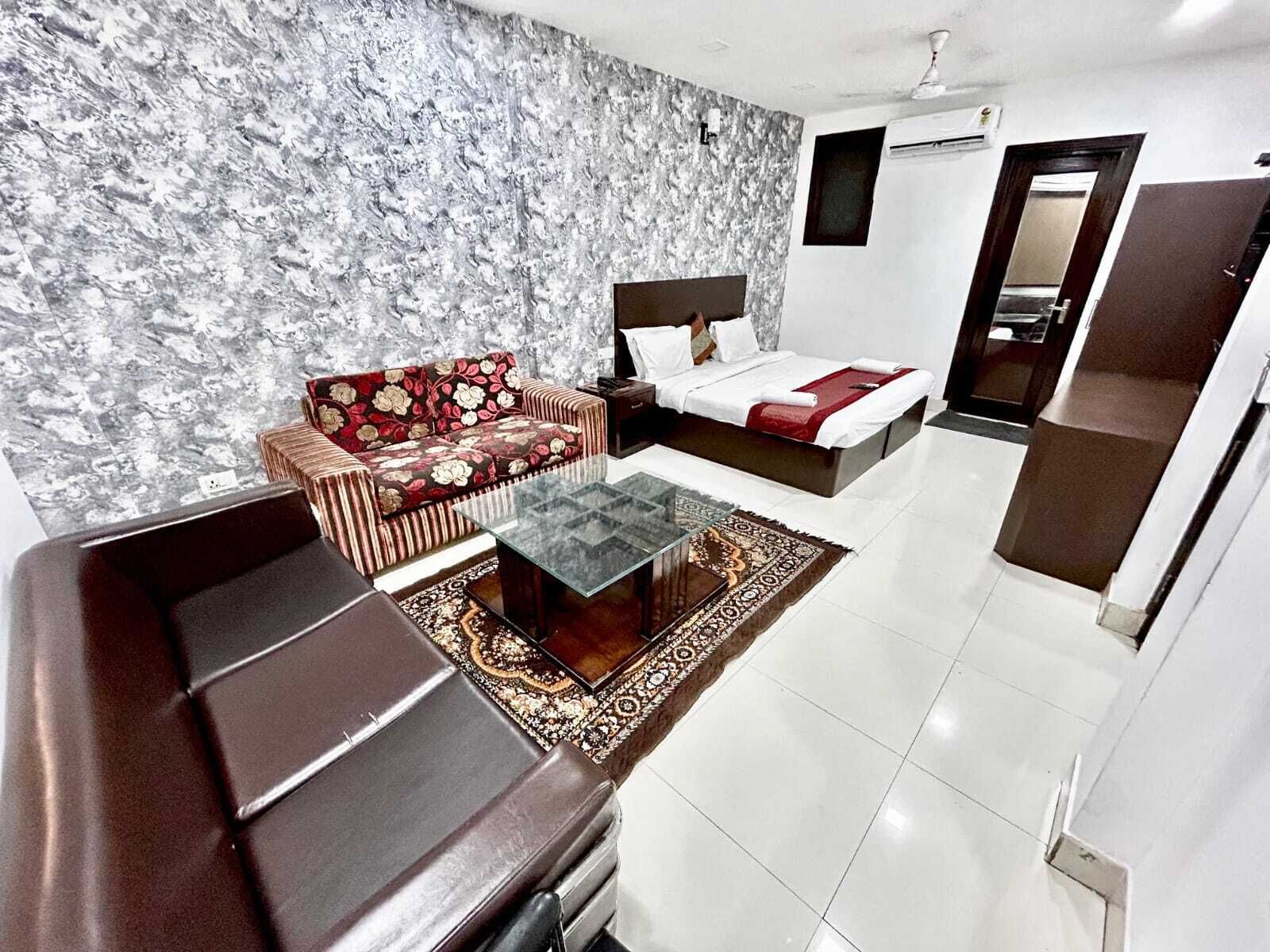 Quarto de hotel moderno com sofá, cama, mesa de centro de vidro e tapete estampado.